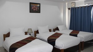 Quarto de hotel com três camas de solteiro, uma mesa de cabeceira com abajur, um quadro na parede e uma janela com cortinas azuis.