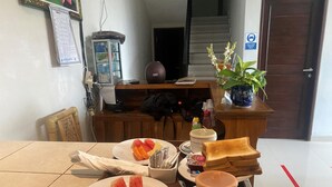 Uma mesa de jantar com pratos de fruta, torradas e uma pequena tigela de sopa. Há uma planta em uma mesinha lateral e uma porta com uma placa ao fundo.