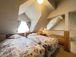 Quarto com duas camas, janela com cortinas, mesa de cabeceira e cômoda de madeira.