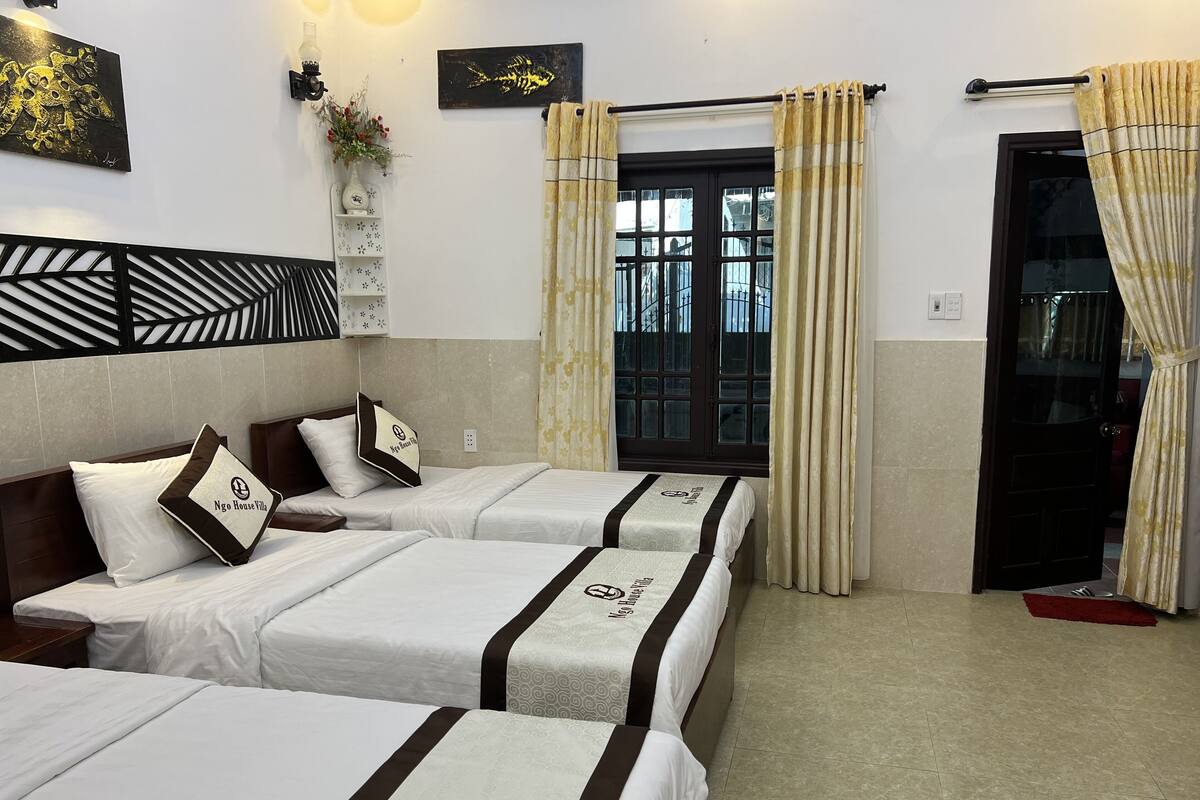Quarto de hotel com três camas, uma janela com cortinas, uma prateleira na parede com um vaso, e uma porta que dá para outro quarto.