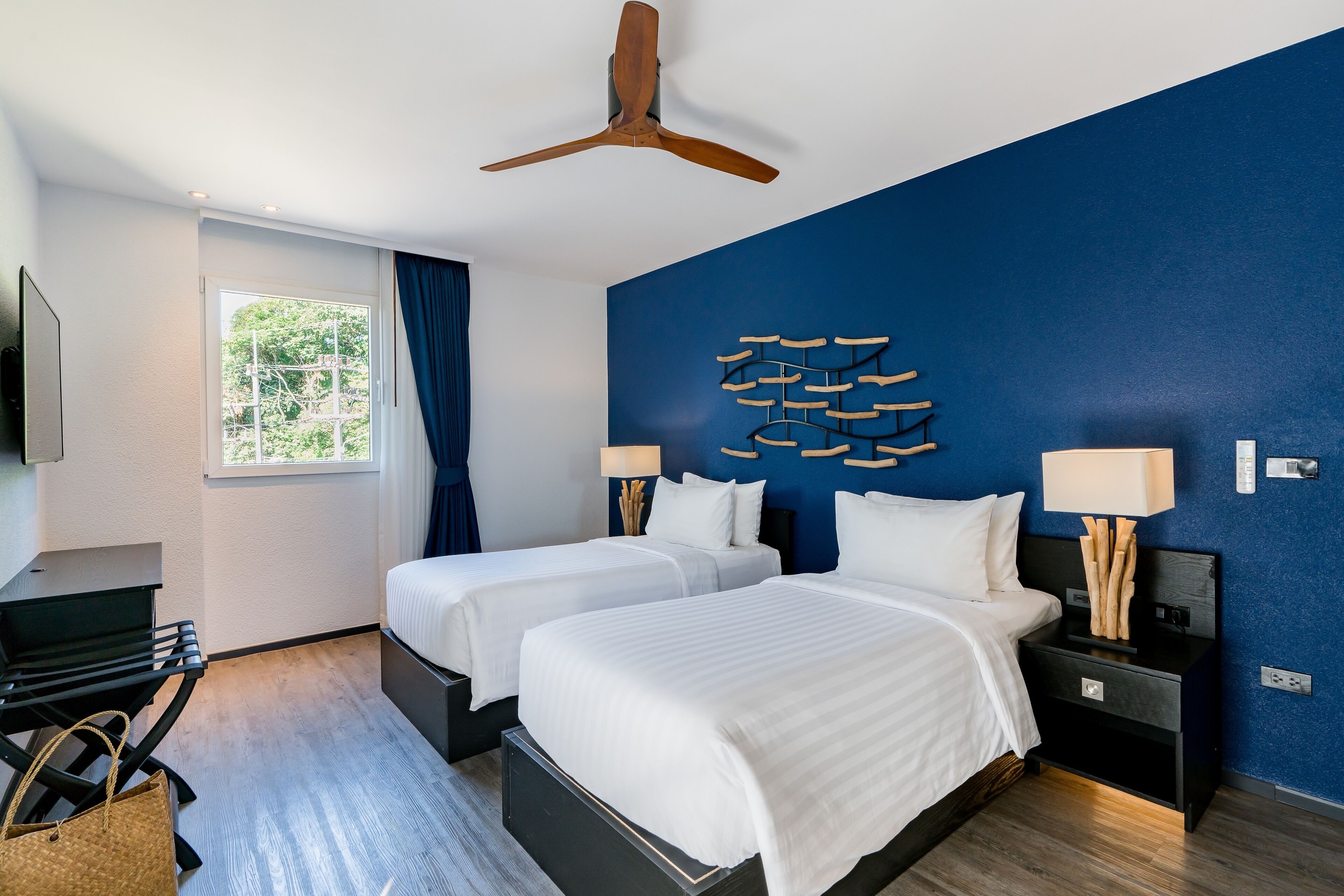 Quarto de hotel com duas camas, uma escrivaninha, uma cadeira, televisão, ventoinha no teto e janela com cortinas.