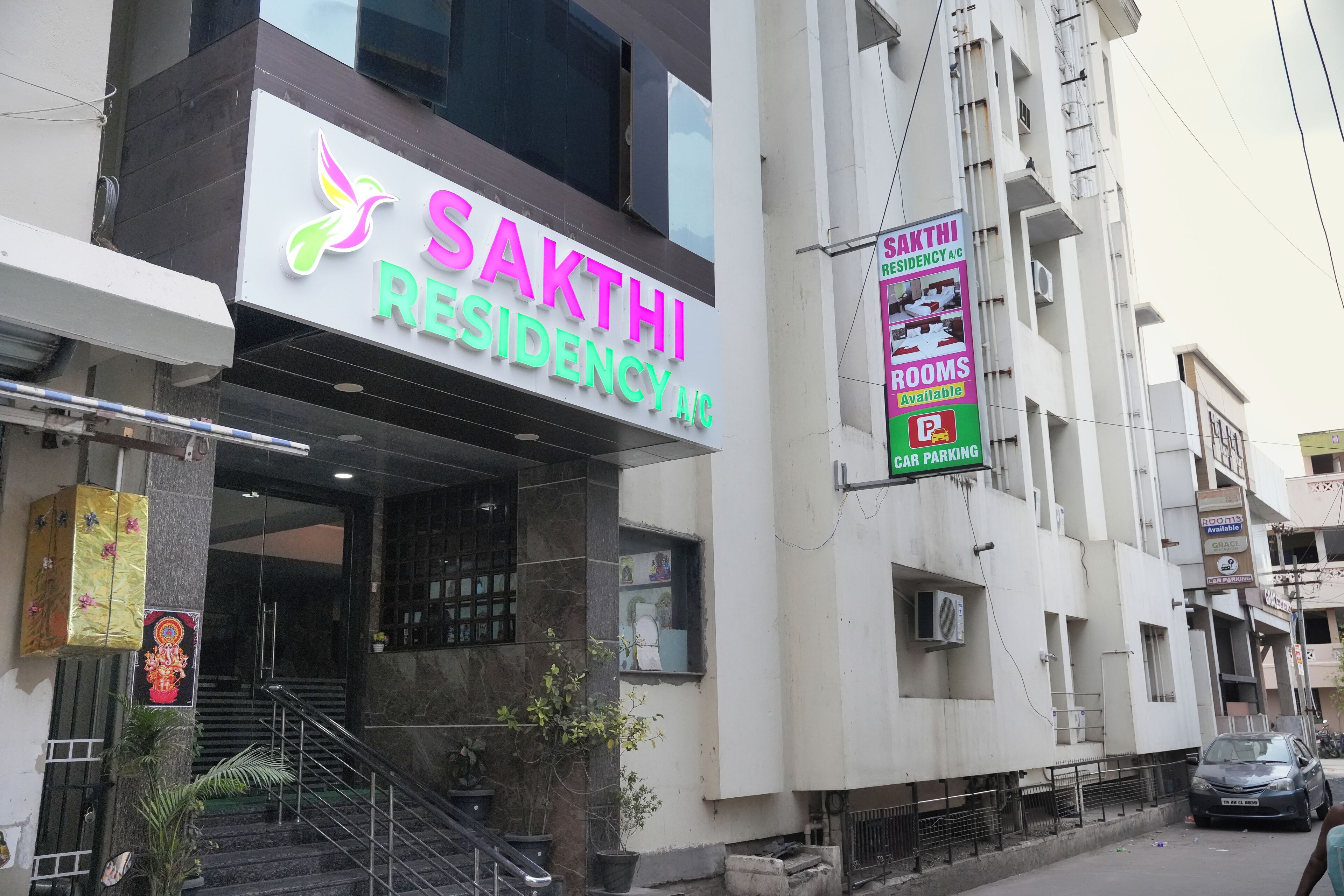 Um edifício com uma placa indicando 'SAKTHI RESIDENCY A/C', com quartos disponíveis e estacionamento para carros.
