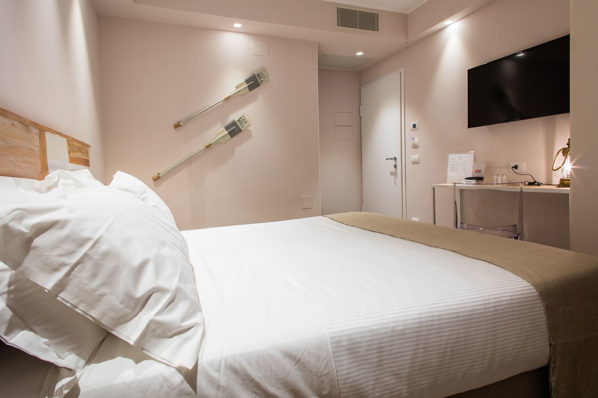 Quarto de hotel com cama, televisão presa na parede e secretária com computador e uma chávena.