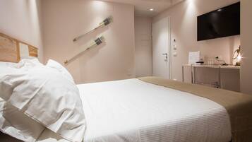 Quarto de hotel com cama, televisão presa na parede e secretária com computador e uma chávena.
