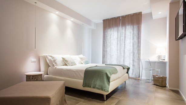 Quarto moderno com cama, mesa de cabeceira e pequena escrivaninha com abajur.