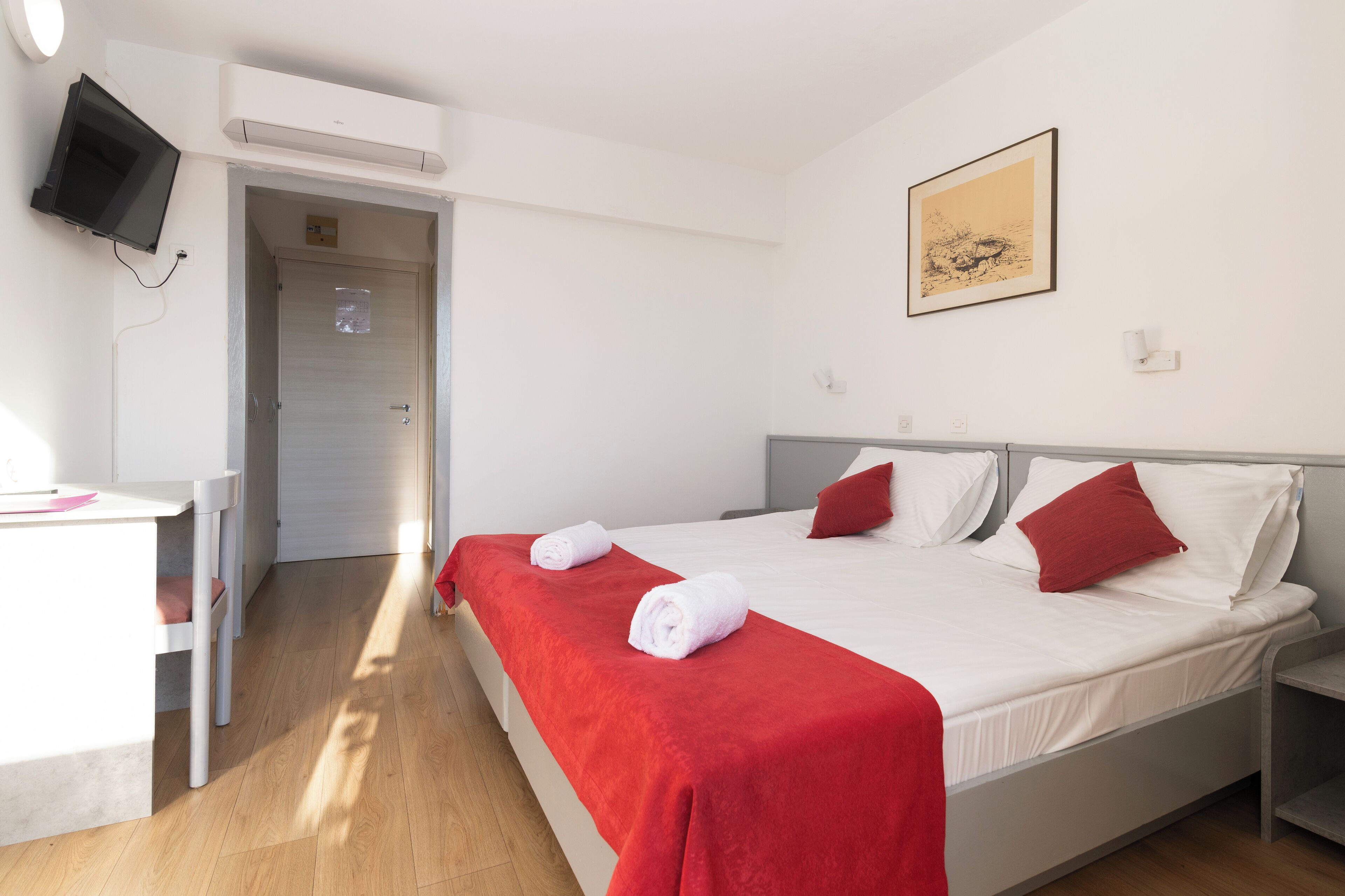 Quarto de hotel com uma cama, escrivaninha, cadeira, televisão e porta.