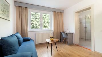 Sala moderna com sofá azul, uma pequena mesa de madeira com um vaso, box de vidro para o chuveiro e vista para o verde lá fora.