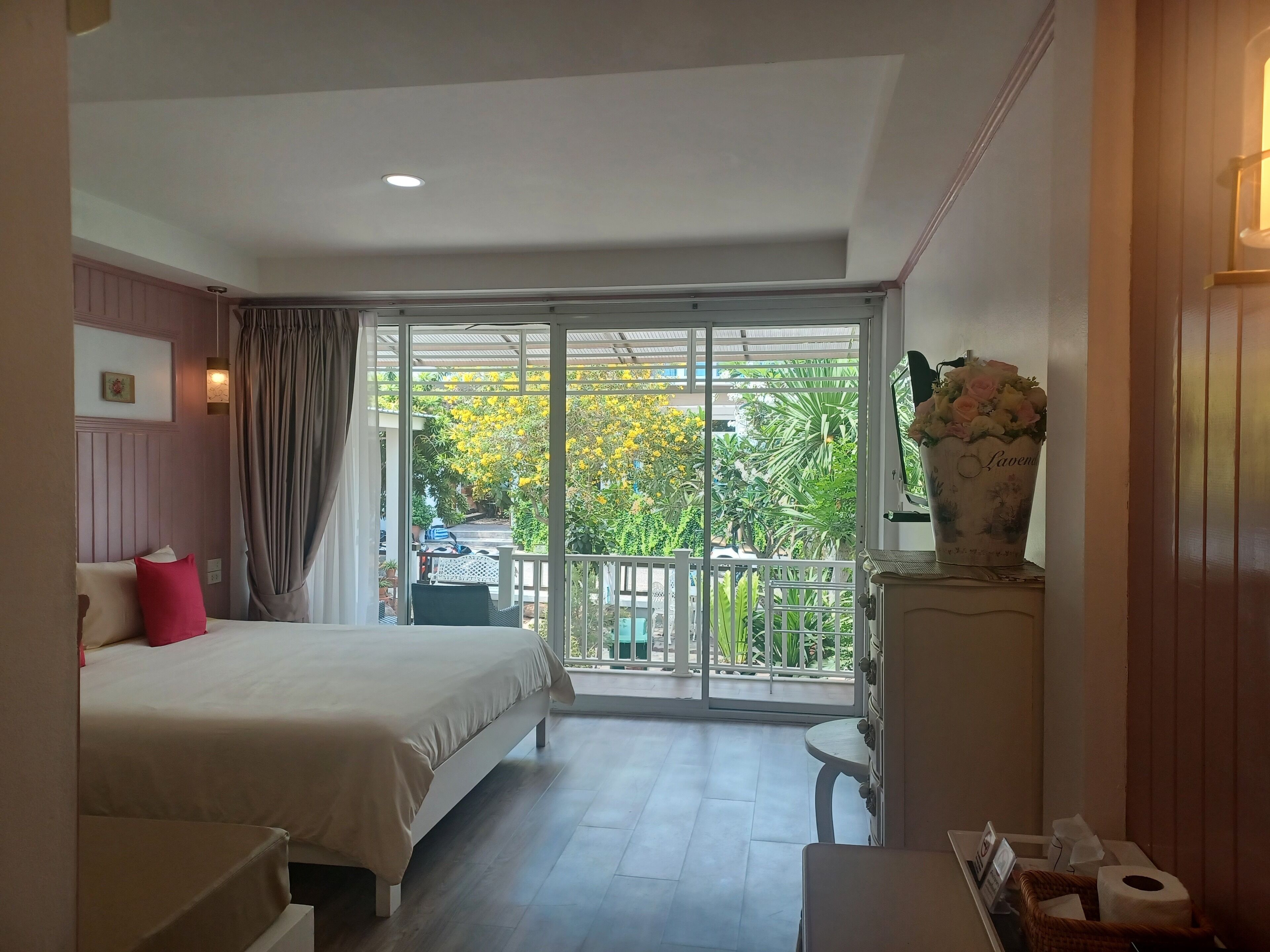 Quarto de hotel com cama, mesa pequena, um vaso de flores e varanda com vista para vegetação e flores amarelas.