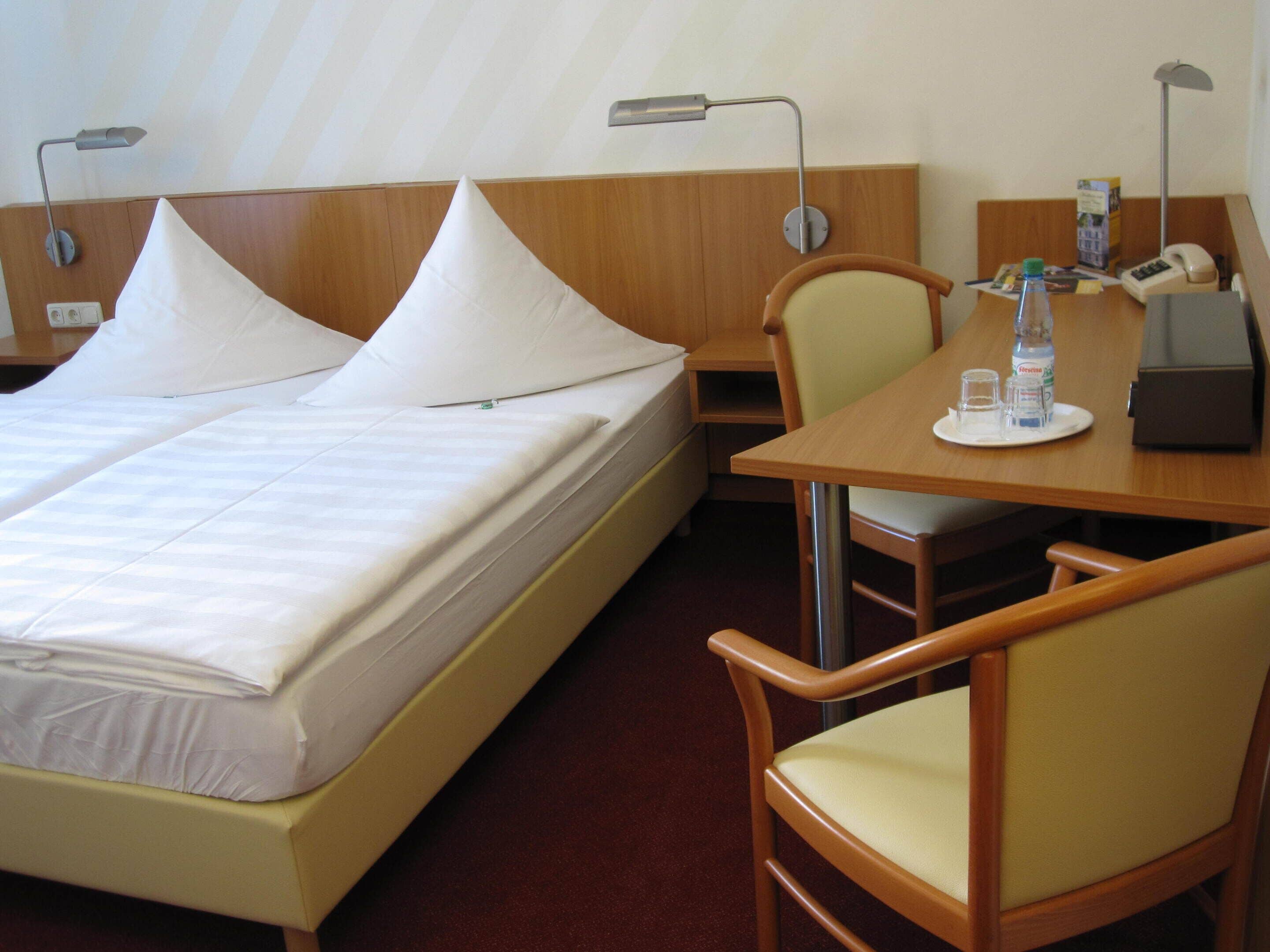 Quarto de hotel com duas camas, uma escrivaninha com cadeira, telefone e uma garrafa de água.