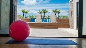 Porta aberta que dá para uma varanda com uma bola de exercícios vermelha, duas cadeiras de descanso e uma piscina ao fundo.