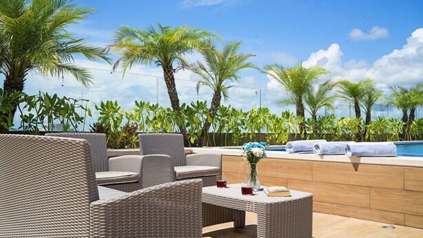 Uma área de estar no terraço com móveis de vime, piscina e palmeiras.