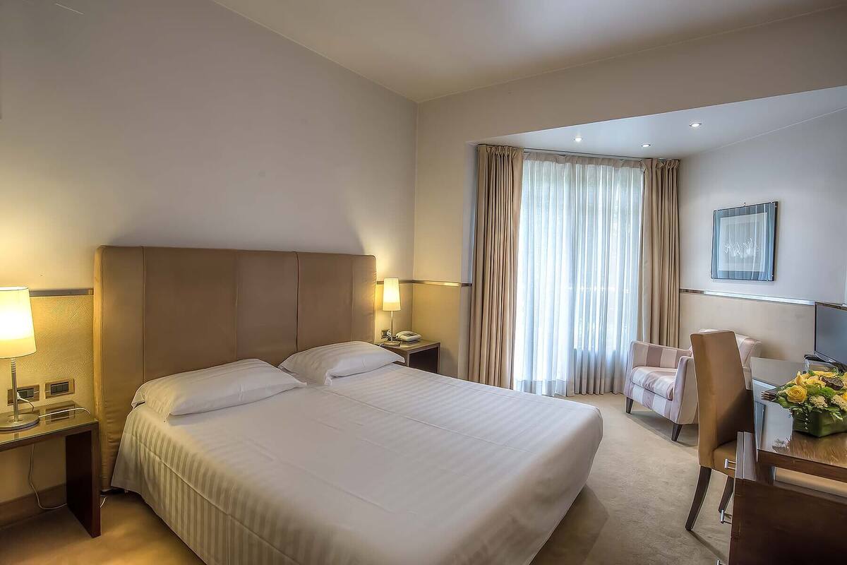 Quarto de hotel com uma cama grande, luminárias de cabeceira, uma escrivaninha com cadeira e uma janela com cortinas.