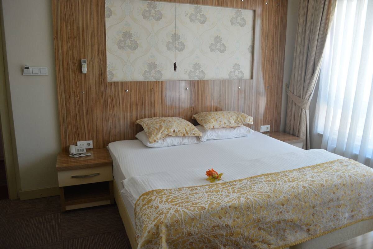 Quarto de hotel com cabeceira de madeira, cama grande com roupa de cama branca, duas mesas de cabeceira e uma televisão fixada na parede.
