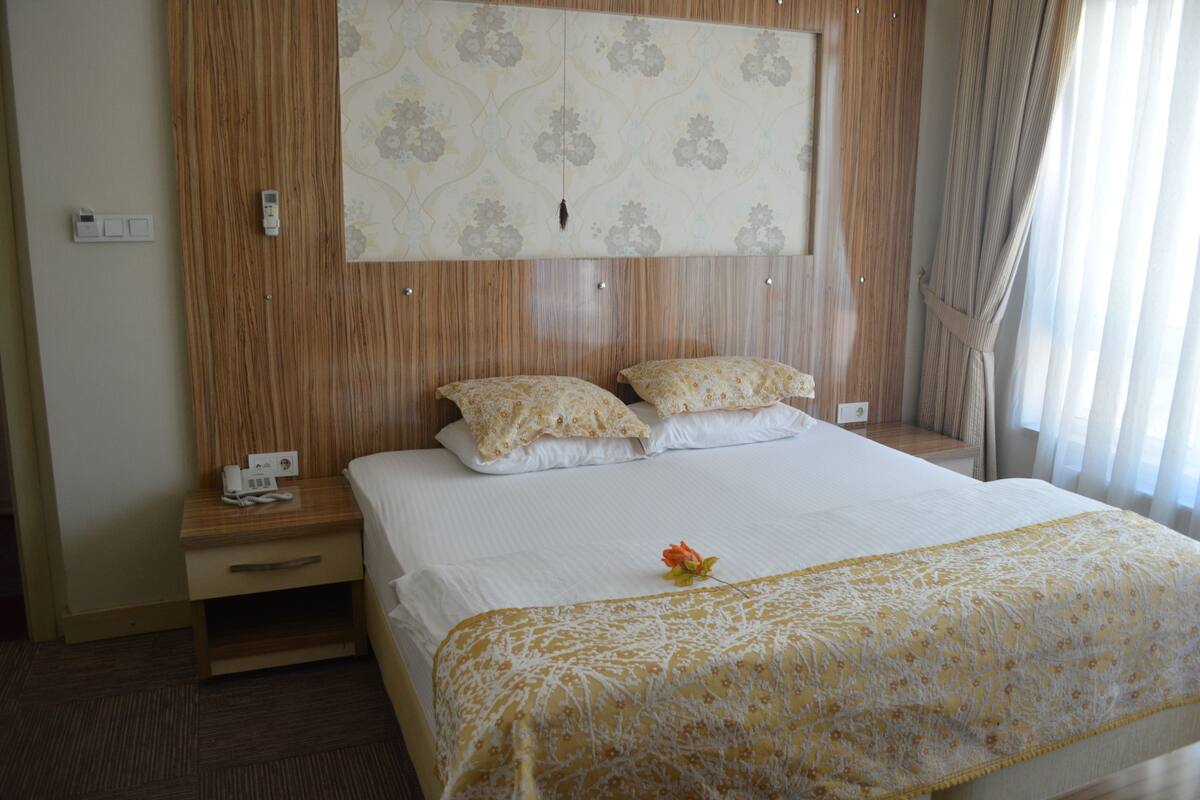 Quarto de hotel com cabeceira de madeira, cama com roupa de cama branca, mesa de cabeceira com telefone e abajur, e janela com cortinas.