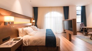 Quarto de hotel com cama, luminárias laterais, televisão de tela plana, escrivaninha com cadeira e janela com cortinas.