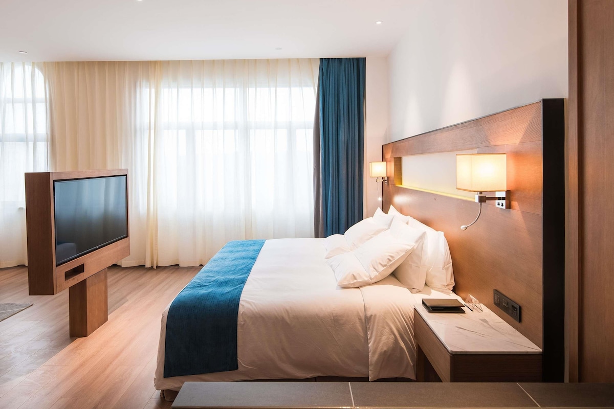Quarto de hotel com cama, mesa de cabeceira, luminária e televisão.