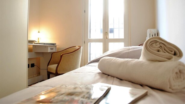 Quarto de hotel com cama, escrivaninha com cadeira, janela com persianas e tomada na parede.