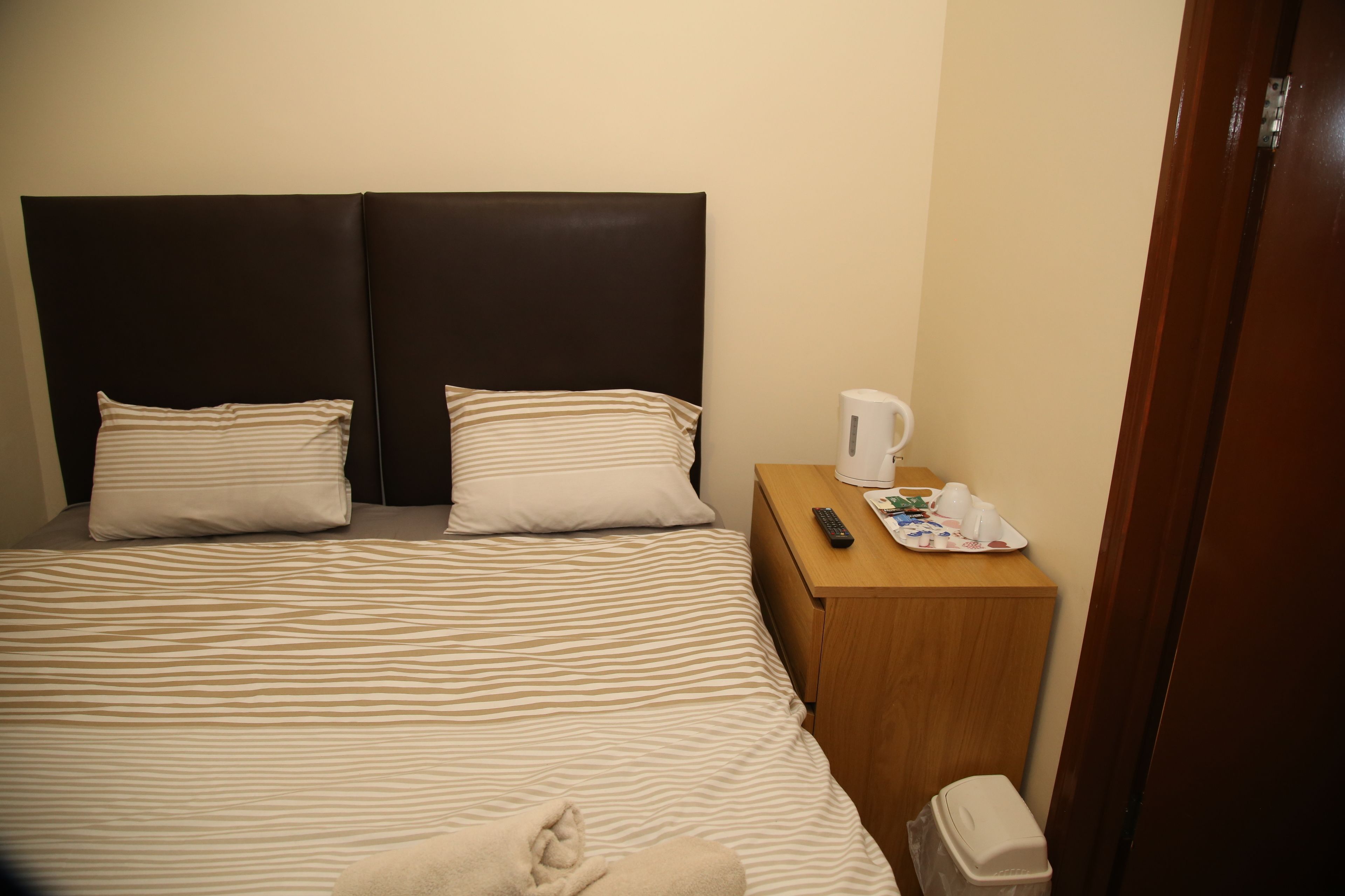 Quarto de hotel com uma cama, roupa de cama listrada, mesa de cabeceira de madeira com um controle remoto, chaleira e uma lixeira.