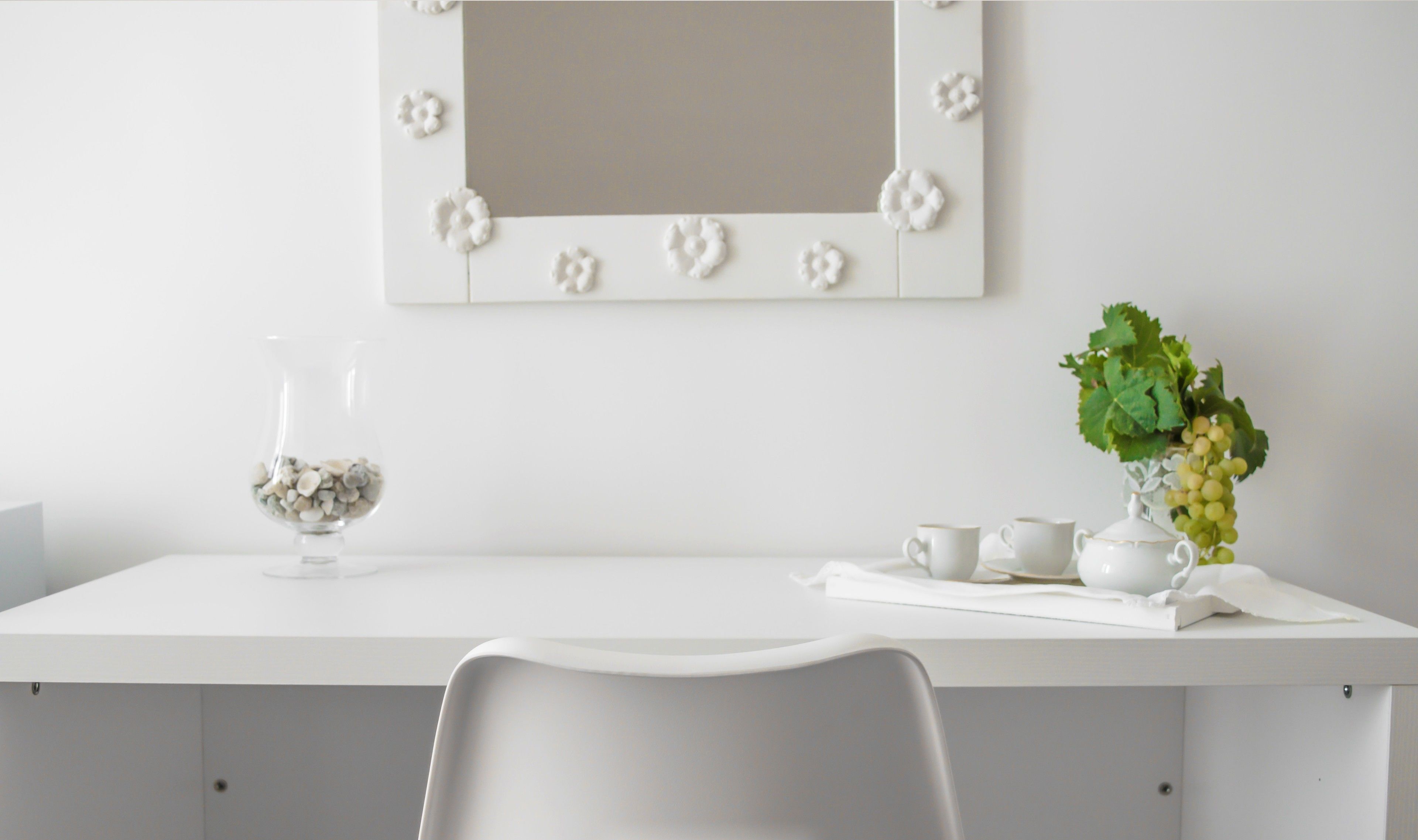 Uma mesa branca com um vaso de uvas verdes e uma taça de vidro transparente com pedrinhas.