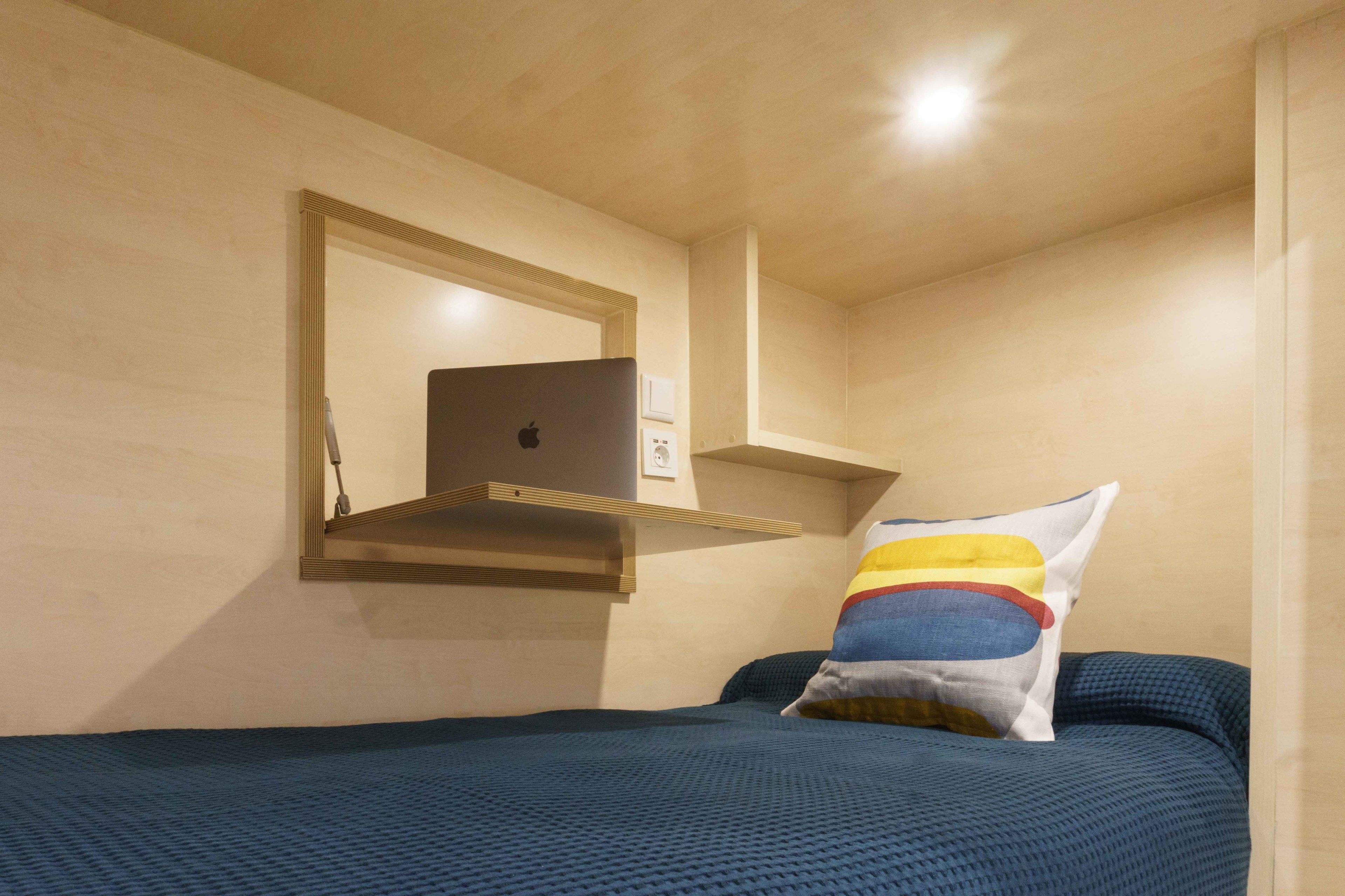 Quarto com cama, um laptop numa estante e um travesseiro listrado.