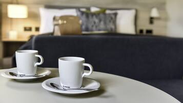Quarto de hotel com uma cama, duas chávenas brancas num pires com uma colher, uma mesa de cabeceira com uma lâmpada e uma parede com um almofado decorativo.