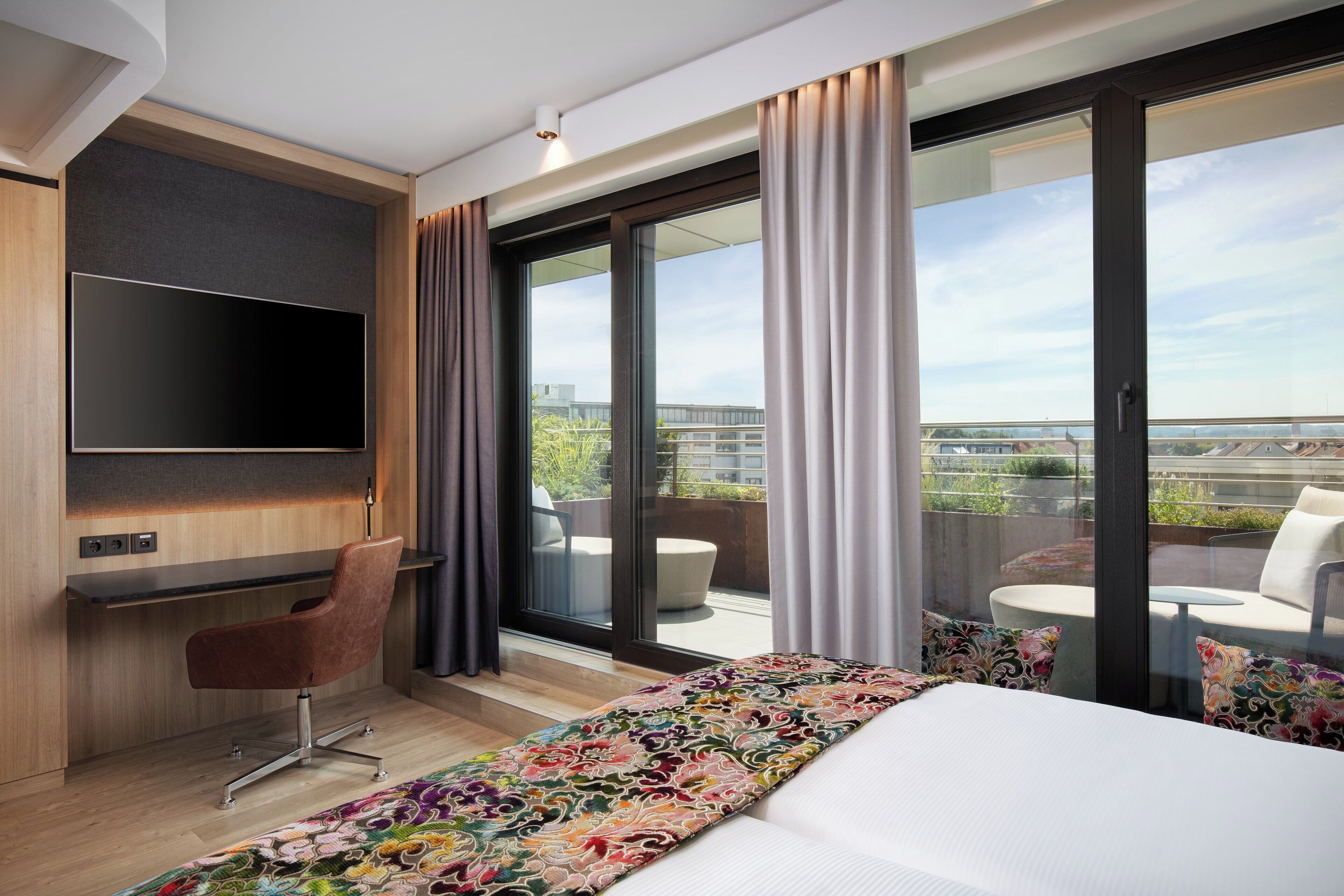 Quarto de hotel moderno com televisão de tela plana, escrivaninha, cadeira, cama e varanda com vista para a cidade.