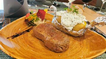 Prato com bife, dois acompanhamentos embalados em papel alumínio e molhos, e um copo de vinho tinto.