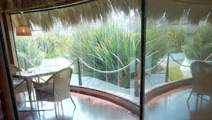 Quarto com parede de vidro que dá para um jardim, uma cadeira de vime, uma mesa pequena e uma toalha no chão.