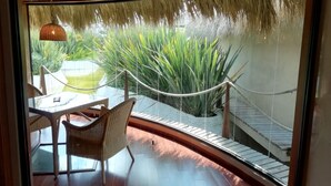 Quarto com uma grande janela de vidro que dá para um jardim com telhado de palha, um deck de madeira e uma área de jantar com móveis de vime.