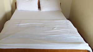 Cama de casal com roupa de cama branca e cabeceira de madeira num quarto com paredes simples.