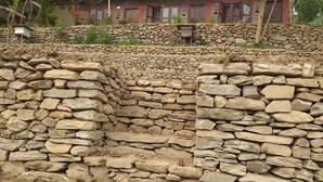 Estrutura de parede de pedra com um banco de pedra e uma pequena mesa, rodeada por vegetação e um edifício com detalhes em vermelho ao fundo.