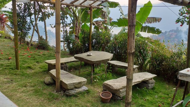 Um jardim com mesas e bancos de pedra, rodeado por vegetação exuberante e vista para uma cidade ao longe.
