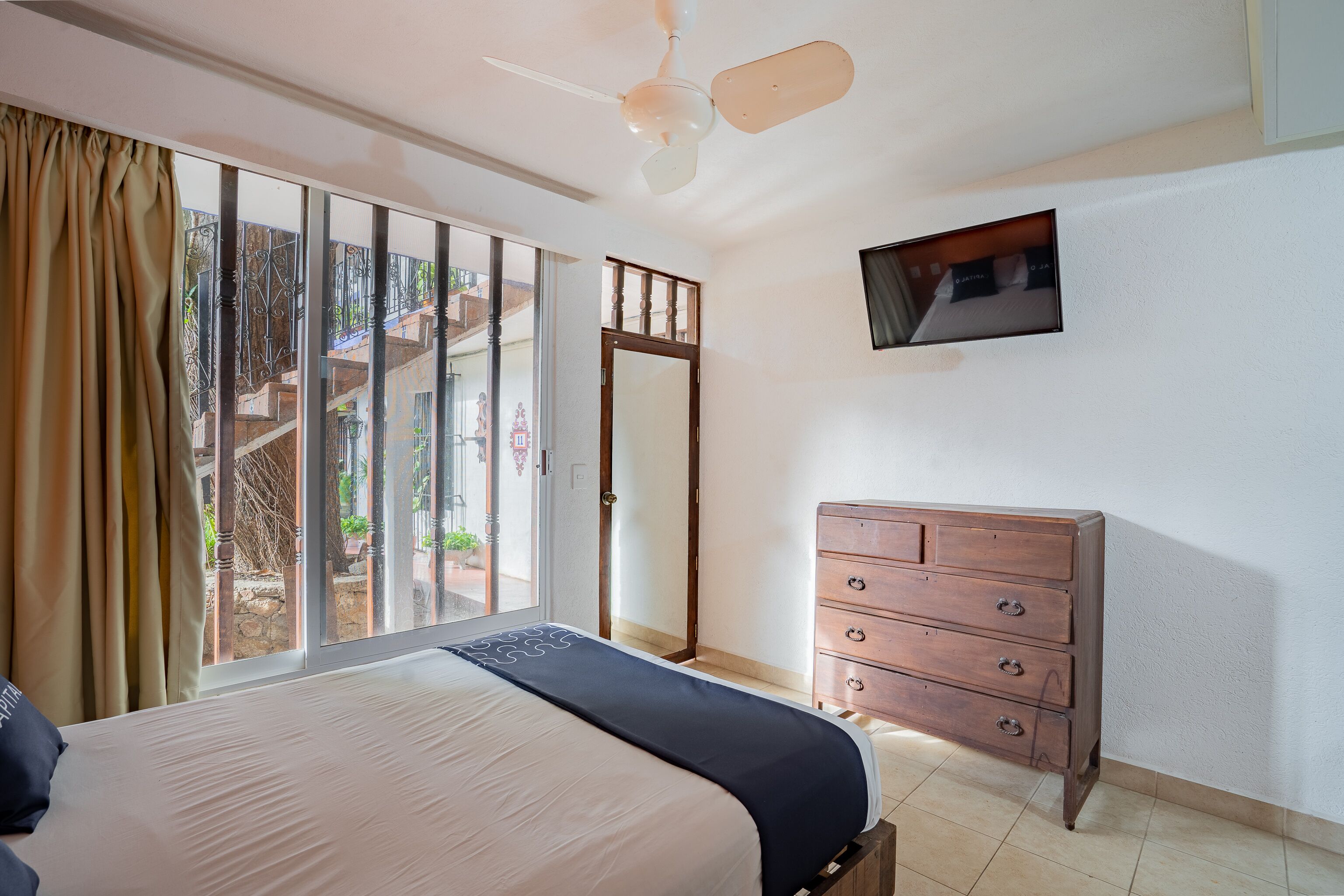Quarto com cama, penteadeira, ventilador de teto, televisão e porta de vidro corrediça que dá para uma varanda.
