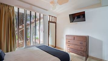Quarto com cama, penteadeira, ventilador de teto, televisão e porta de vidro corrediça que dá para uma varanda.