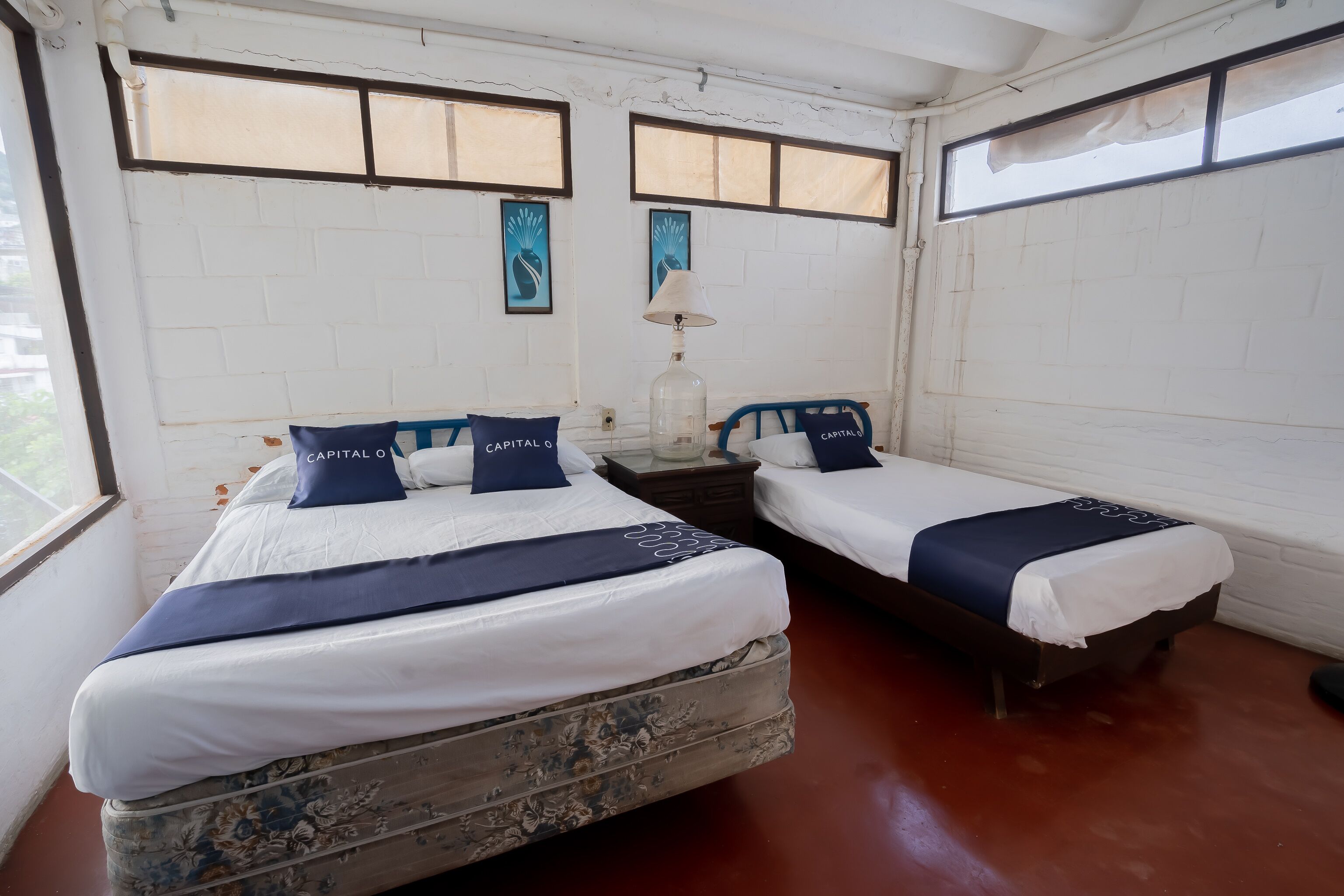 Quarto com duas camas, cada uma com colchão branco e cobertor azul. Um criado-mudo com abajur fica entre as camas. As paredes do quarto são de tijolos brancos e há uma janela com vista para o exterior.