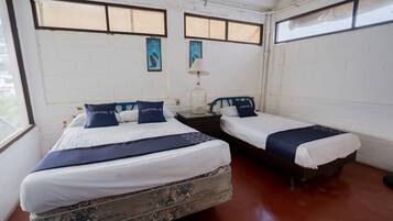 Quarto com duas camas, cada uma com colchão branco e cobertor azul. Um criado-mudo com abajur fica entre as camas. As paredes do quarto são de tijolos brancos e há uma janela com vista para o exterior.
