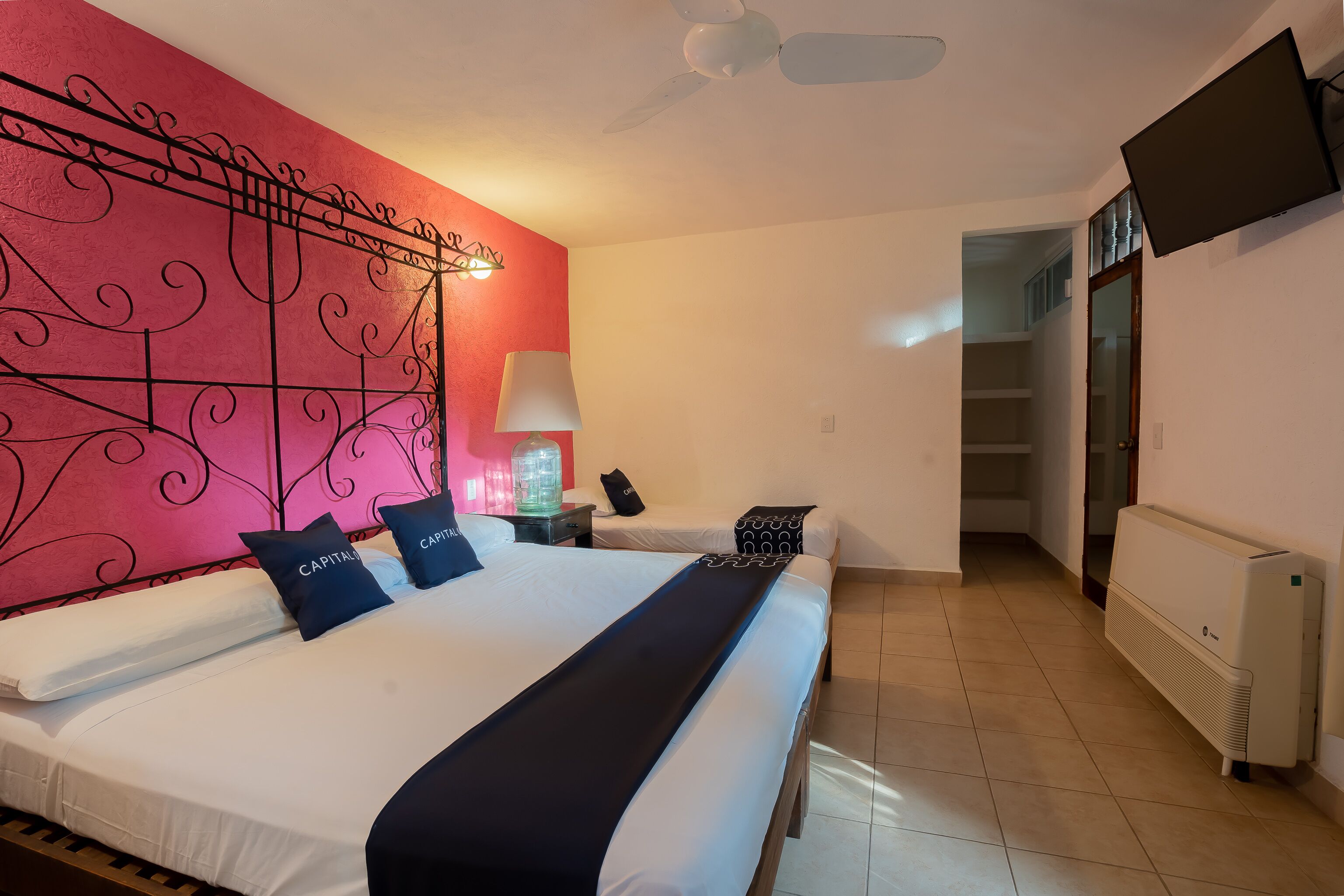 Quarto de hotel com uma cama grande, um ventilador de teto, uma TV presa na parede e um dispensador de água na mesa de cabeceira.