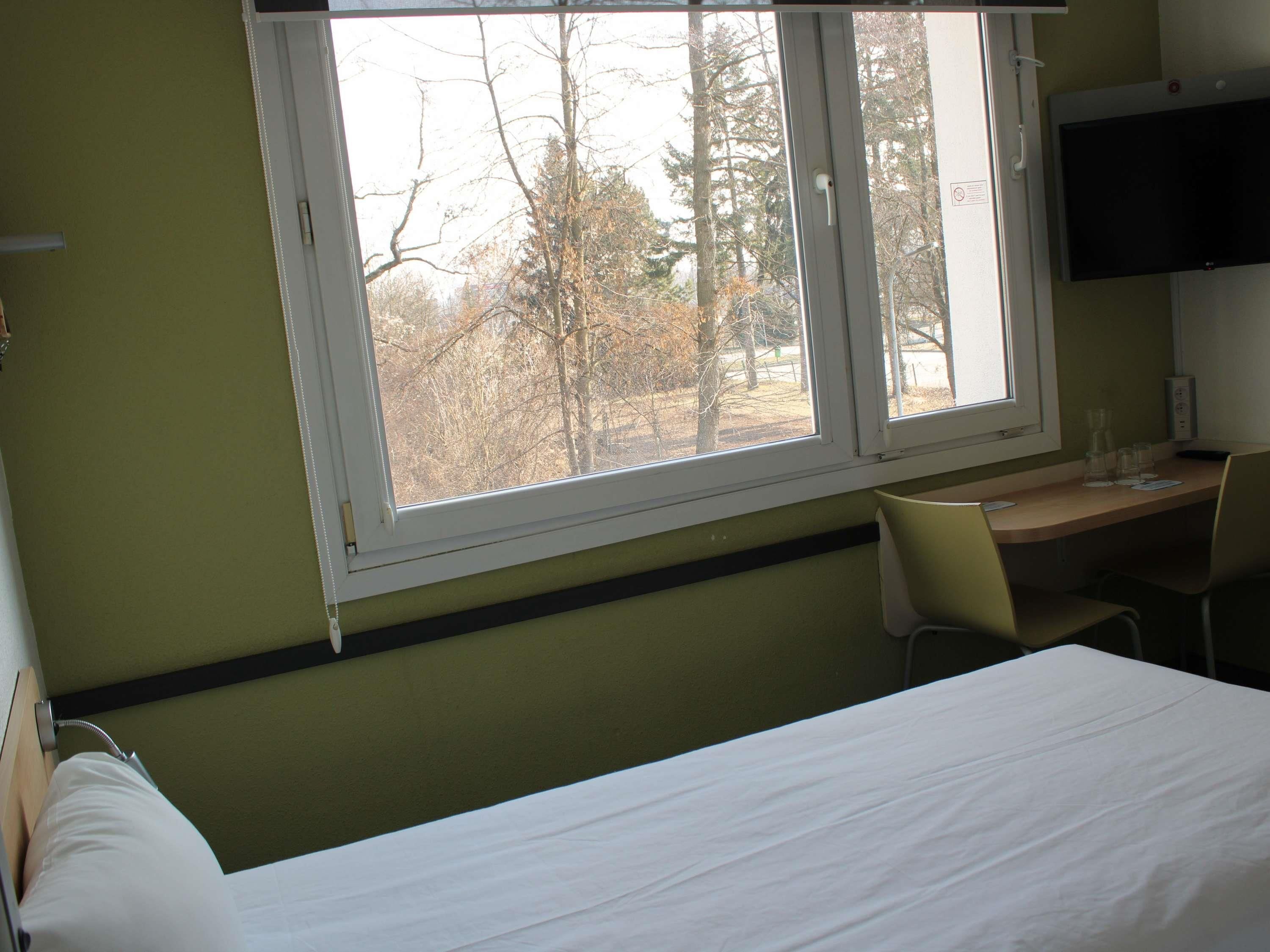Quarto de hotel com cama, escrivaninha e cadeira. Janela com vista para árvores e um edifício.