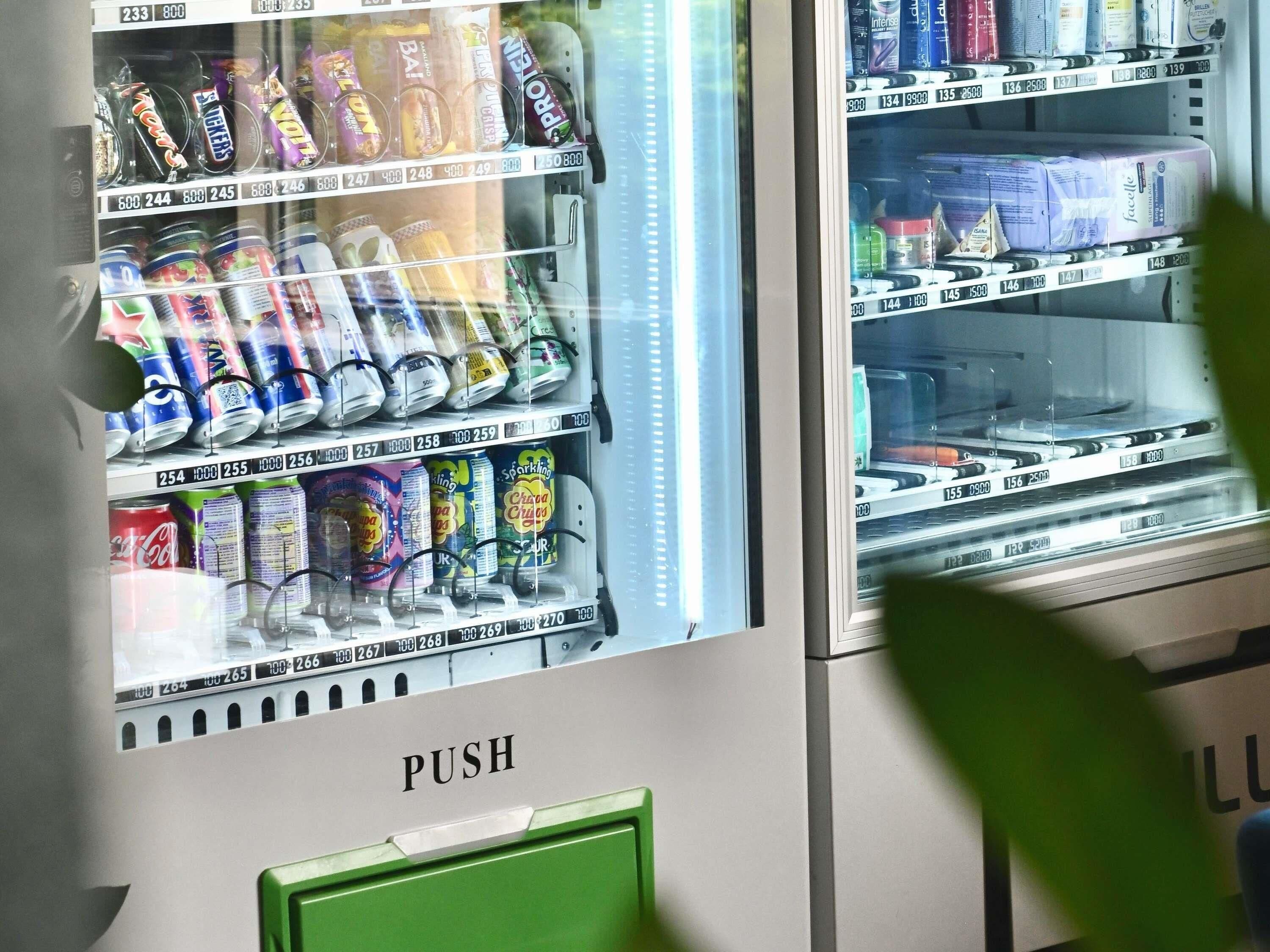 Máquina de vendas com lanches, bebidas, um botão verde e uma prateleira com produtos de higiene pessoal.