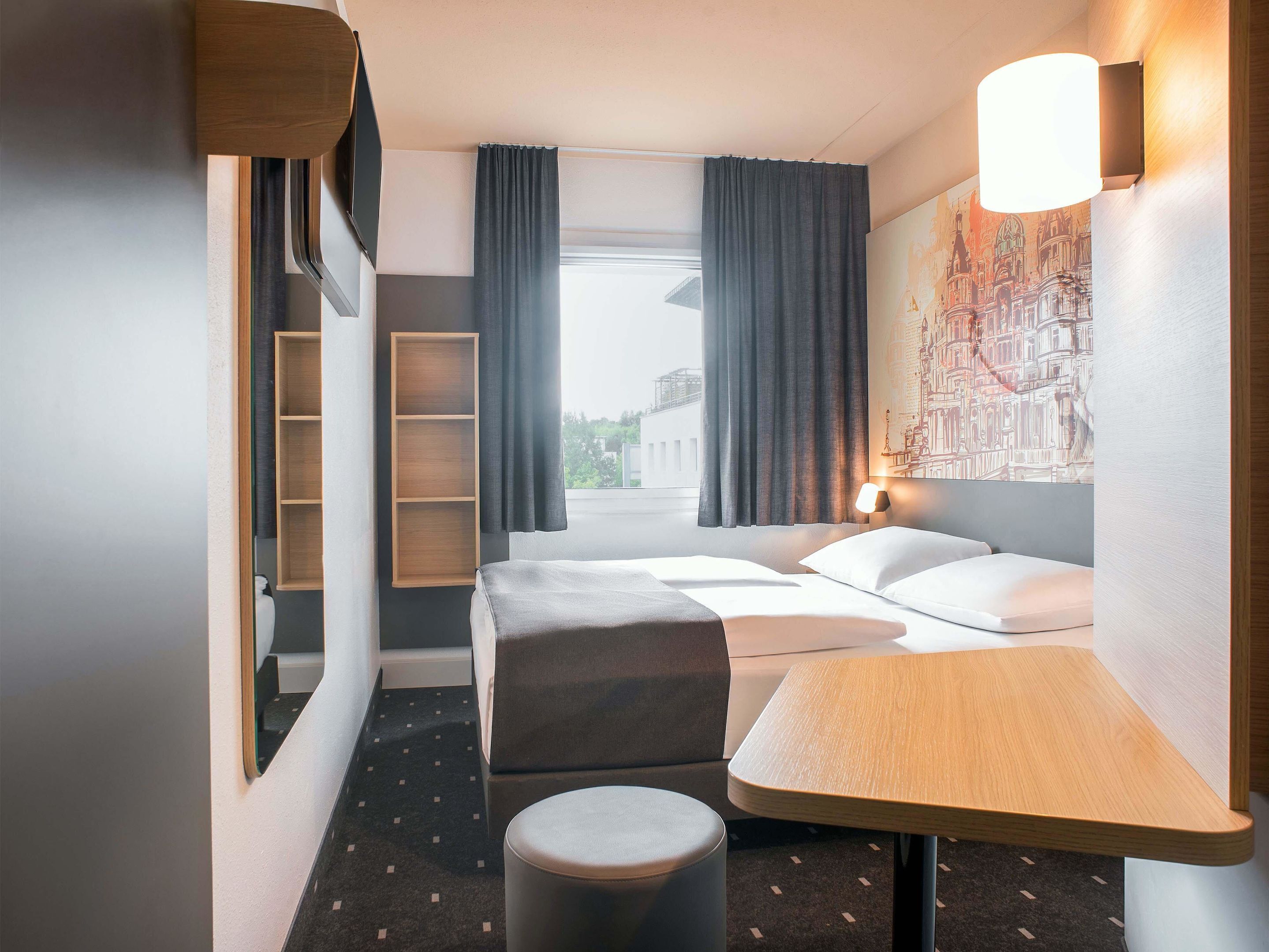 Quarto de hotel com cama, uma pequena mesa, cadeira, prateleira e um quadro na parede.