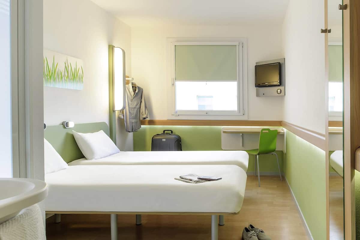 Quarto de hotel com duas camas, uma escrivaninha, uma cadeira, uma televisão e uma mala.