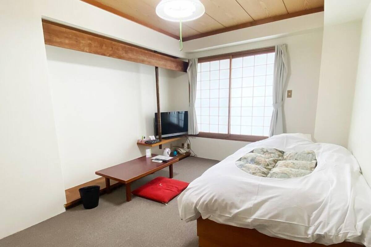 Quarto com uma cama, TV, escrivaninha de madeira e janela.