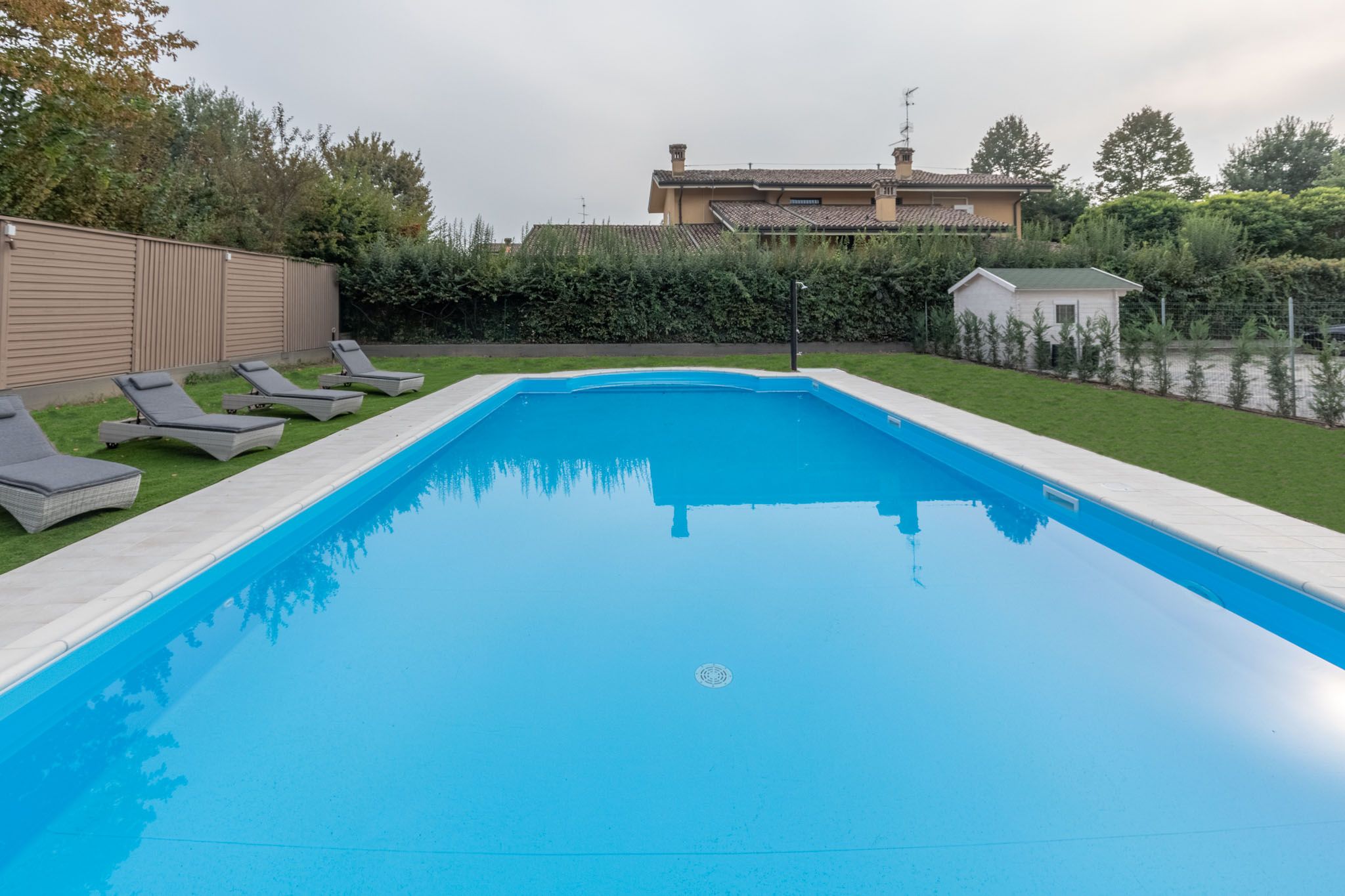 Uma piscina retangular com superfície azul límpida, rodeada por um deck revestido com azulejos. Três cadeiras de descanso estão dispostas ao longo da borda da piscina.
