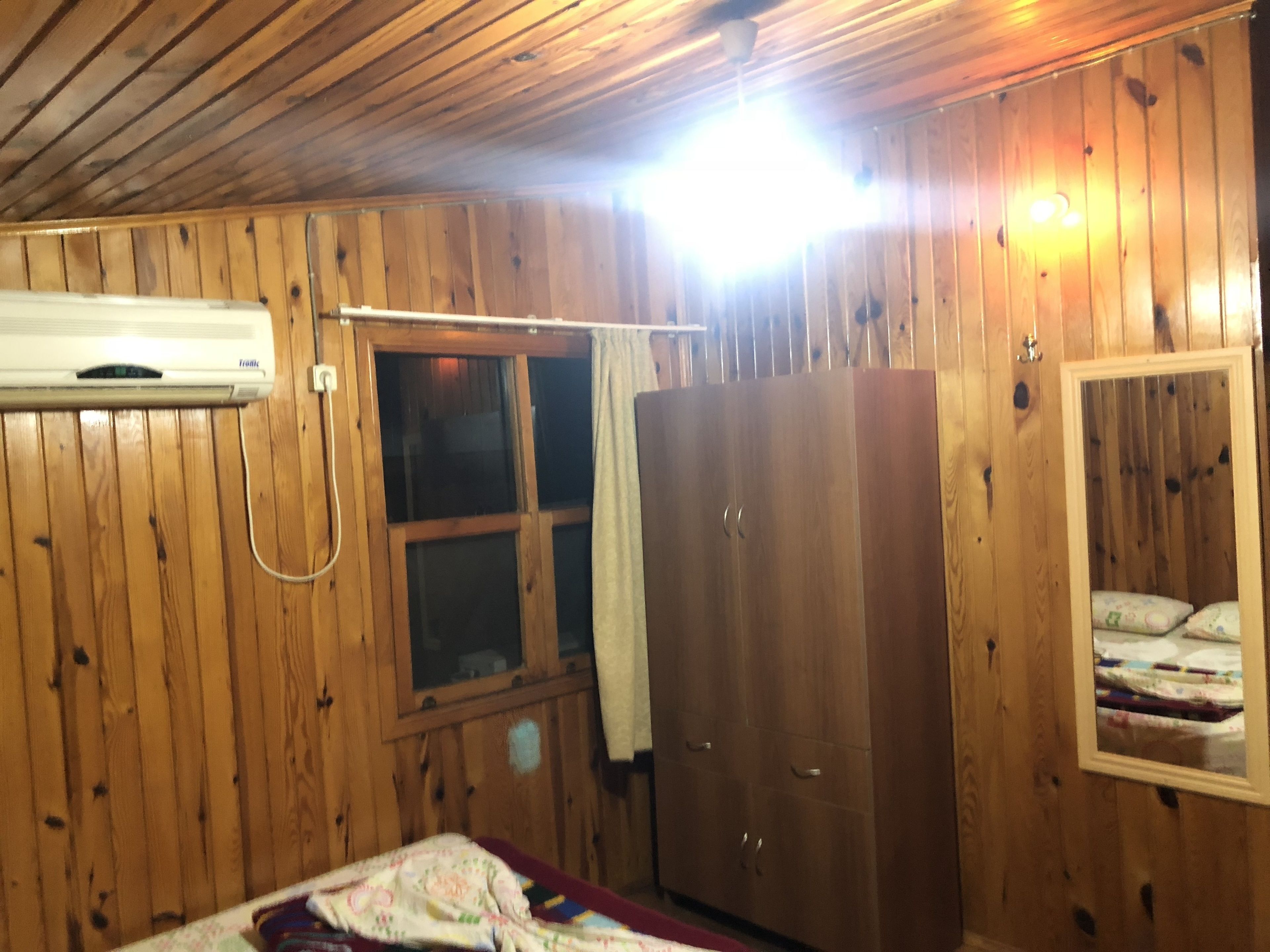 Quarto com painéis de madeira, uma cama, um guarda-roupa, uma janela com cortinas e um aparelho de ar condicionado.