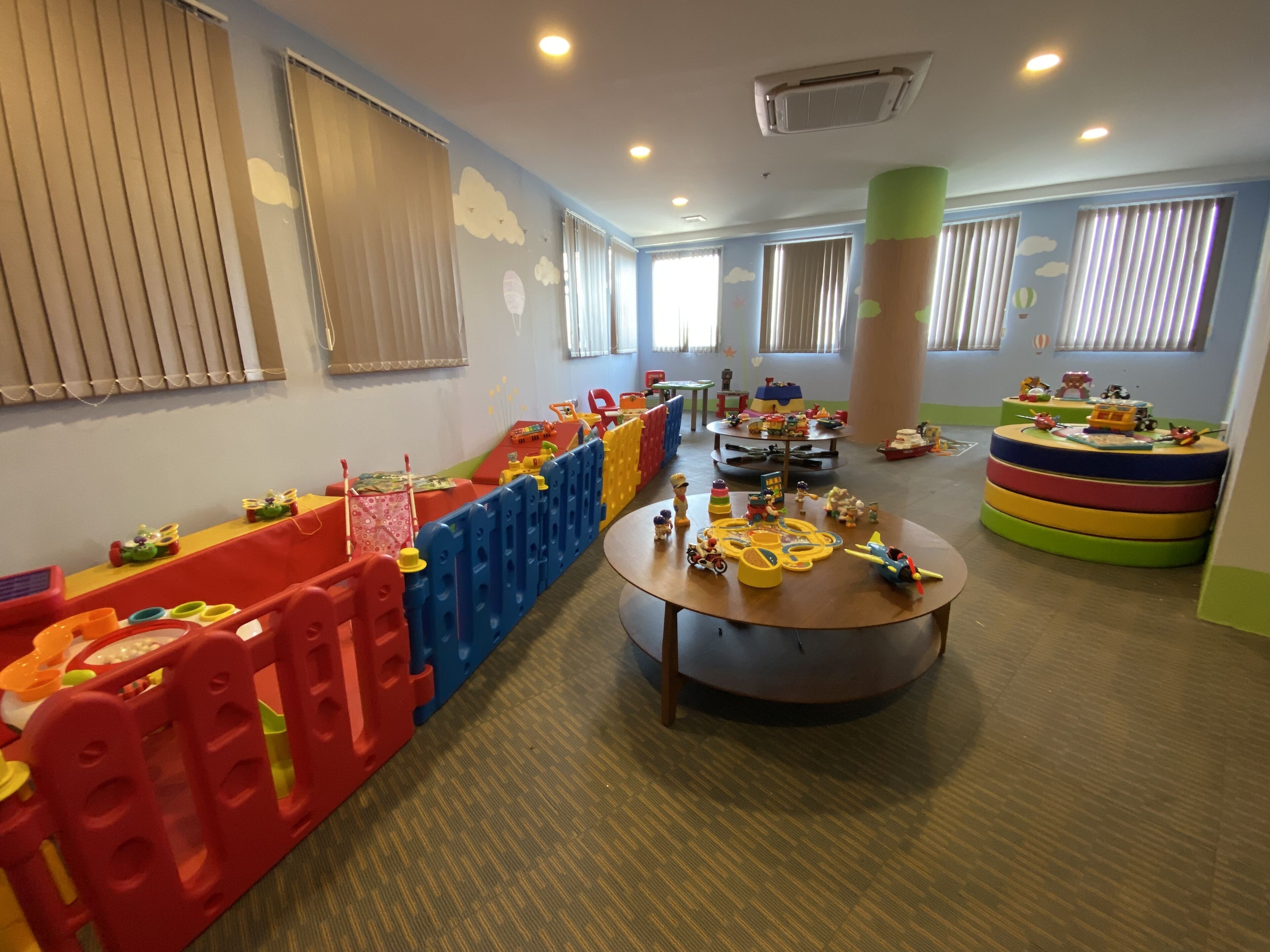 Uma área de recreação com estruturas coloridas, uma mesa central com brinquedos e amplas janelas com cortinas.