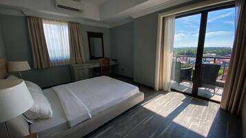 Quarto de hotel com cama, escrivaninha, cadeira, uma janela grande com vista para prédios e um ar condicionado.