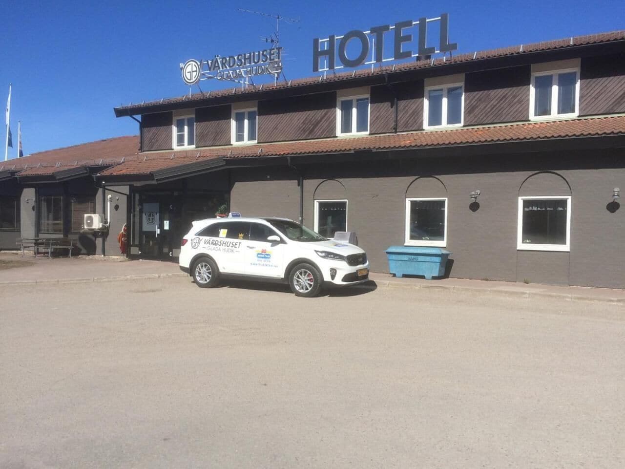 Um hotel com um carro branco estacionado em frente, uma placa com a inscrição "VARDSHUSET" e um contentor de lixo azul à direita.