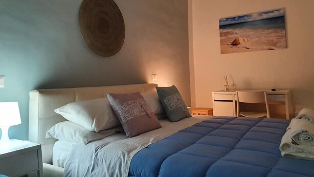 Um quarto com uma cama, mesas de cabeceira, um criado-mudo, uma luminária na parede e uma pintura de uma praia.