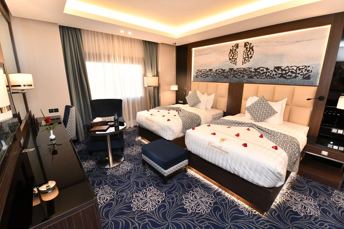 Quarto de hotel com duas camas, uma escrivaninha, uma cadeira, uma televisão e uma grande obra de arte na parede.