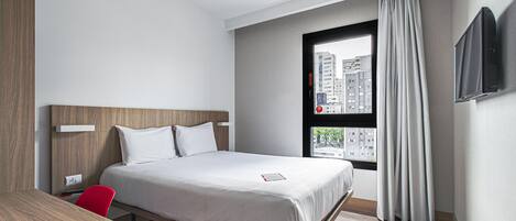 Quarto de hotel moderno com uma cama, uma escrivaninha, uma cadeira vermelha, uma televisão e uma janela com vista para a cidade.